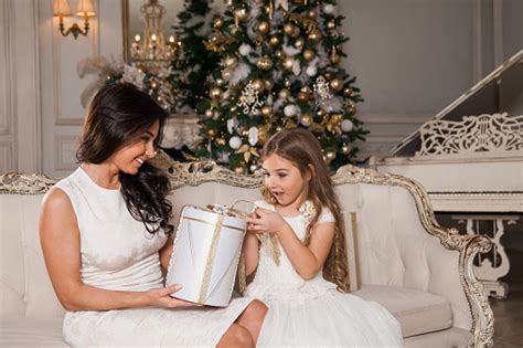 메리 크리스마스와 해피 홀리데이 쾌활한 엄마와 그녀의 귀여운 딸 소녀는 피아노와 장식 된 크리스마스 트리의 배경에 흰색 고전적인 인테리어에서 선물을 교환합니다 새해 2021