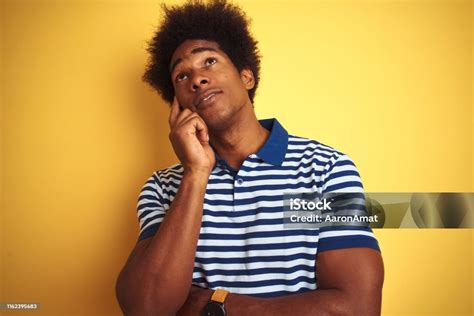 해군 줄무늬 폴로를 입고 고립 된 노란색 배경 위에 서있는 아프로 머리를 가진 미국 남자는 턱에 손을 얹고 질문 펜시브 표현을 생각하고 있습니다 사려 깊은 얼굴로 미소 의심