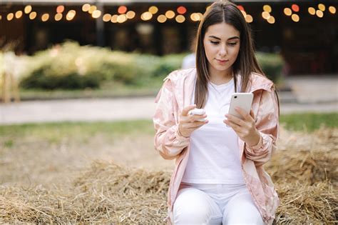 매력적인 젊은 여성은 그녀의 휴대 전화에 위테리스 헤드폰을 원뿔하려고합니다 카페 야외에서 건초 벤치에 앉아있는 여성 Brand Name Smart Phone에 대한 스톡