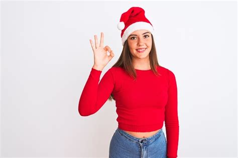 고립 된 흰색 배경 위에 서있는 크리스마스 산타 모자를 쓰고있는 어린 아름다운 소녀는 손과 손가락으로 확인 기호를하고 긍정적 인 미소를 짓고 있습니다 성공적인 식입니다 12