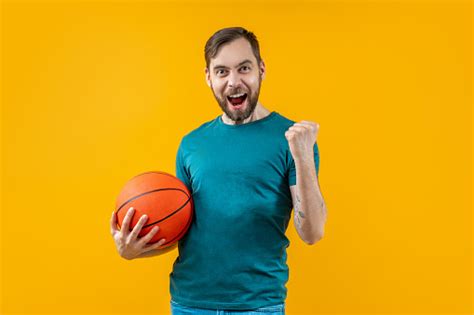 밝은 색상의 주황색 노란색 배경 위에 포즈를 취하고 공을 손에 들고 우승자의 제스처가 주먹을 움켜쥐는 농구 팬의 초상화 남자에
