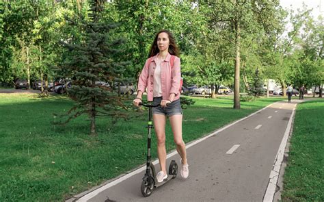 분홍색 재킷을 입은 소녀는 여름에 도시에서 스쿠터 배경 도로 및 녹색 잔디밭을 타고 길을 인도합니다 여성 데님 반바지 텍스트 복사본을 위한 여유 공간입니다 녹색 나무와 나무