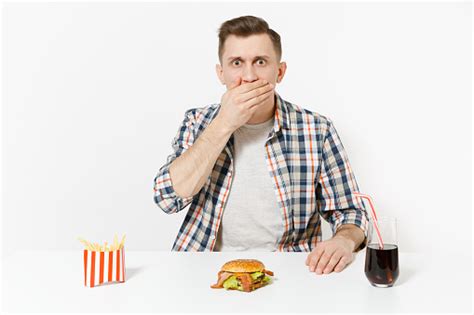 젊은 남자와 햄버거 감자 튀김 콜라 흰색 배경에 고립 된 유리에 테이블에 앉아 손으로 덮개 입 적절 한 영양 섭취 또는 고전적인 미국의 패스트 푸드입니다 광고 공간을 복사