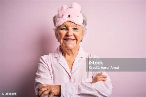 수면 마스크와 잠옷을 입고 고립 된 분홍색 배경 행복한 얼굴을 입고 카메라를 바라보는 교차 된 팔로 미소짓는 선임 아름다운 여성 긍정적 인 사람 긍정적인 감정 표현에 대한