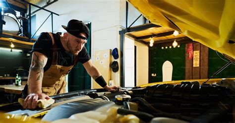 앞치마와 야구 모자를 쓴 젊은 진지한 남자가 자동차의 열린 후드 위로 몸을 구부리고 차고나 작업장에서 엔진 점검을 하고 있다