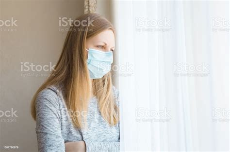 입과 코를 가리는 인공 호흡기 마스크를 착용 한 젊은 성인 여성은 흰색 커튼에 서서 창문을 들여다 봅니다 바이러스 시간에 가정