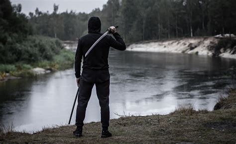 뒷모습 검은 옷을 입은 한 남자가 어깨에 카타나를 들고 강물 근처의 숲에서 카메라를 등지고 서 있습니다 카타나를 가진 용감하고 강한 남자 가공의 인물에 대한 스톡 사진 및