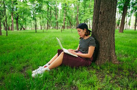 캐주얼 한 옷과 스냅 백 야구 모자를 입은 어린 소녀는 줄무늬 티셔츠와 긴 버건디 스커트와 흰색 운동화를 녹색 숲의 나무 아래에 흰색 노트북으로 앉아 있습니다 30 34세에