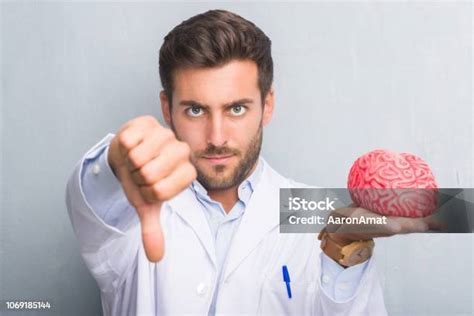 성 난 얼굴 아래로 엄지 싫어 보여주는 음수 부호 거부 개념 뇌를 들고 회색 그런 지 벽에 잘생긴 젊은 의사 심리학자 남자 개념에 대한 스톡 사진 및 기타 이미지 Istock
