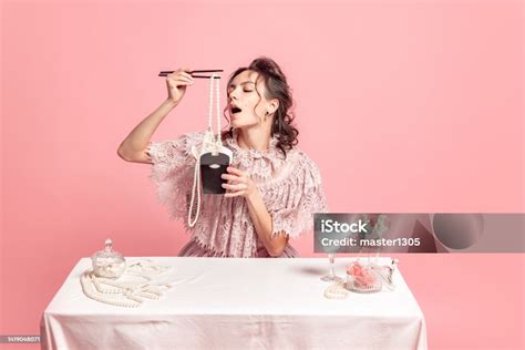 복고풍 스타일의 헤어도를 가진 젊은 아름다운 여성이 분홍색 배경에 고립 된 서빙 테이블에 앉아있는 중세 드레스를 입은 창조적 인 이미지 30 34세에 대한 스톡 사진 및