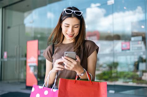 패션 몰에서 휴대 전화를 사용하여 쇼핑백으로 아름다운 여성을 즐기십시오 트렌디 한 여성이 옷 가게 창문 근처에 스마트 폰을 들고 있습니다 쇼핑 라이프 스타일 패션을 즐기는