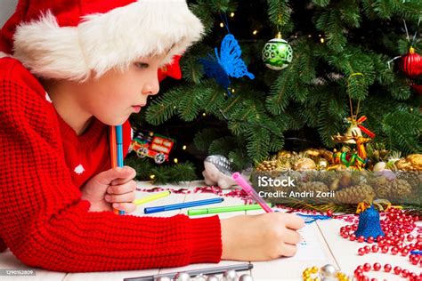 어린 아이는 산타에게 편지를 씁니다 산타 모자를 쓴 귀여운 소년이 크리스마스 트리 근처에서 산타에게 편지를 씁니다 겨울에 대한