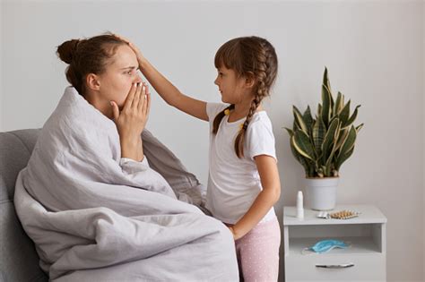 소파에 앉아 아픈 건강에 해로운 여자의 실내 샷 담요에 싸여 손바닥으로 입을 덮고 그녀의 매력적인 배려 딸은 엄마의 이마를 만지고 온도를 측정 25 29세에 대한 스톡 사진