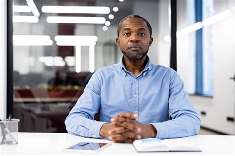 현대적인 사무실의 책상에 앉아 두 손을 깍지 낀 자신감 넘치는 사업가는 결단력과 집중력을 반영합니다 책상 위에 펼쳐진 책과 스마트폰이 있는 전문적인 분위기 남자에 대한 스톡