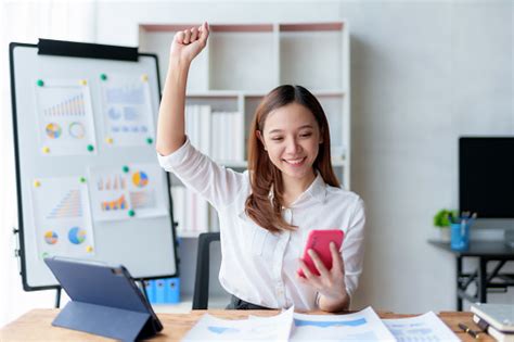 스마트 폰을 사용하는 아름다운 아시아 사업가는 사무실 휴대 전화를 통해 달성 한 성공에 기뻐하고 흥분합니다 Brand Name Smart Phone에 대한 스톡 사진 및