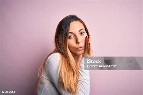 젊은 아름다운 금발의 여자 와 파란 눈 입고 흰색 티셔츠 위에 분홍색 배경 손 에 입 말하기 비밀 소문 속삭이는 악의적 인 대화 사생활에 대한 스톡 사진 및 기타 이미지