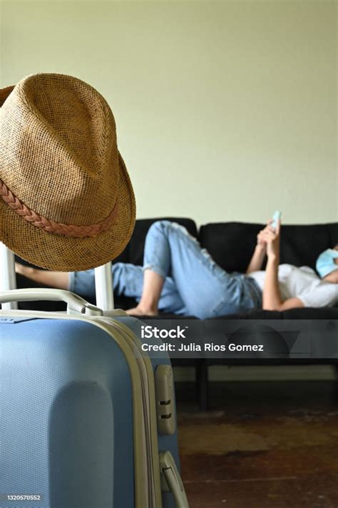 밀짚 모자를 쓴 파란색 가방의 수직 클로즈업 스마트폰을 들고 있는 마스크를 쓰고 검은 색 소파에 누워 있는 청바지를 입고 복사 공간을 가진 젊은 여성 Brand Name