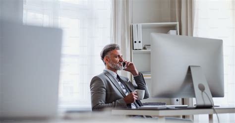 스마트폰 커피 데스크톱 컴퓨터를 가진 진지한 성숙한 사업가가 사무실의 테이블에 앉아 전화를 걸고 있다 사진 Unsplash의 성인 이미지