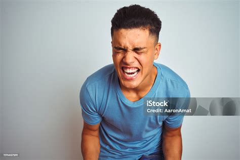 고립 된 흰색 배경 위에 서있는 파란색 티셔츠를 입은 젊은 브라질 남자는 팔을 들고 미소를 지으며 성공을 위해 비명을 지르며 우승자 제스처를 하고 매우 행복하고 흥분했습니다