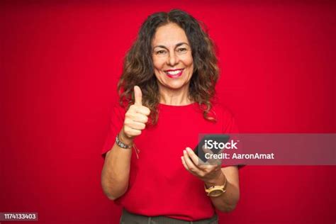 빨강 고립된 배경을 통해 스마트 폰을 사용하는 중년 노인 여성은 큰 미소로 행복한 확인 사인을하고 손가락으로 엄지 손가락 우수한 기호를 사람들에 대한 스톡 사진 및 기타