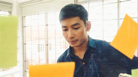 비즈니스 사람들은 사무실의 프로젝트 계획 보드에서 일하고 동료 친구와 대화하여 프로젝트 개발을 분석합니다 그들은 유리 벽에 게시 된 스티커 메모를 사용하여 정리합니다