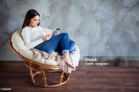 아늑한 베개 의자에 앉아 앉아있는 멋진 매력적인 사랑스러운 사랑스러운 집중 평화로운 꿈갈색 머리 여자는 회색 콘크리트 벽을 통해 흥미로운 에세이 이야기 디지털 전자 책을