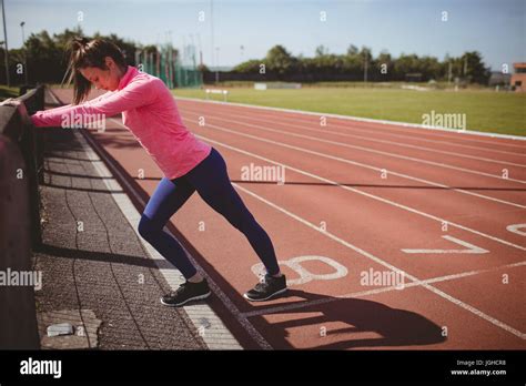 Full Length Of Female Athlete Stretching By Railing On Running Track During Sunny Day Stock