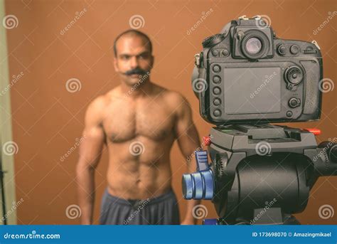 Muscular Indian Man With Mustache Shirtless While Vlogging Against Brown Background Stock Photo