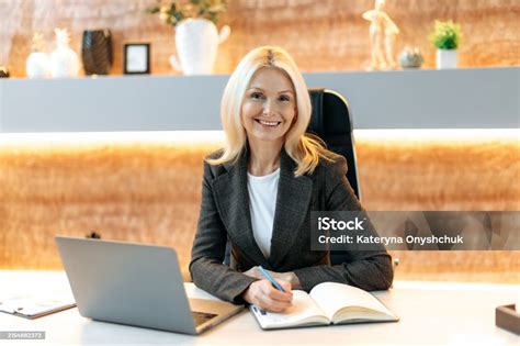 아름 다운 우아한 성공적인 중년 백인 여성 비즈니스 레이디 기업 최고 관리자 정장 사무실 테이블에 앉아 카메라를보고 친절하게 미소 짓는 초상화 45 49세에 대한 스톡 사진