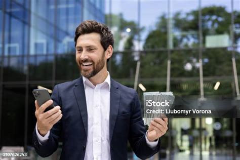 행복한 청년이 달러 지폐 부채를 손에 들고 사무실 근처에 아이돌 슈트를 입고 서서 전화기 화면을 미소 지으며 바라보고 있습니다 35 39세에 대한 스톡 사진 및 기타 이미지