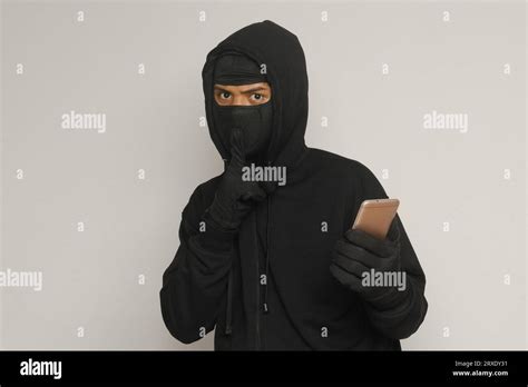 Portrait Of Mysterious Man Wearing Black Hoodie And Mask Doing Hacking Activity On Mobile Phone