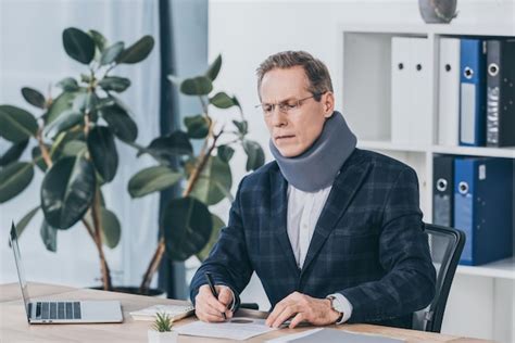 Businessman In Neck Brace Sitting At Table And Writing In Document In