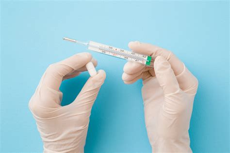 밝은 파란색 테이블 배경에 수은 온도계와 흰색 알약을 들고 고무 보호 장갑에 의사 손 파스텔 색상입니다 근접 촬영 관점 바이러스 시간에 약하를 수신 맨 위 보기 A형