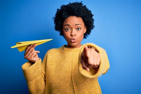 젊은 아프리카계 미국인 여성 곱슬 머리 들고 종이 비행기 위에 파란색 배경 카메라와 당신에 게 손가락으로 가리키는 손 기호 앞에서 긍정적이 고 자신감 제스처 가리키기에 대한