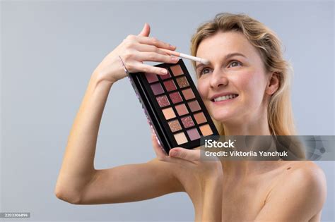 화려한 아이섀도 팔레트를 사용하여 메이크업을 하고 미소를 지으며 거울을 보며 밝은 환경에서 자신의 뷰티 루틴과 셀프 케어 방법을 보여주는 젊은 여성 눈 화장에 대한 스톡