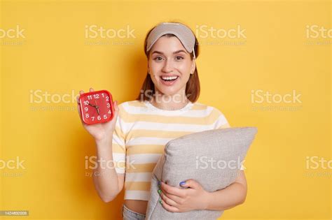 회색 베개와 빨간 알람 시계를 들고 흥분한 카메라를보고 고립 된 노란색 배경을 포즈를 취하는 흥분한 행복한 만족 어두운 머리 여자의 사진 벽 시계에 대한 스톡 사진 및 기타