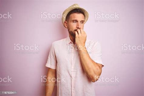 분홍색 고립 된 배경 위에 모자와 여름 셔츠를 입은 잘 생긴 남자는 손톱을 물고 손톱을 물고 손에 스트레스와 긴장 찾고 불안