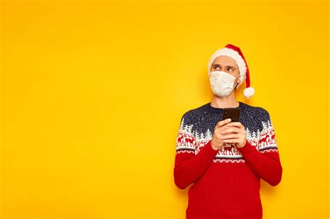새해 옷을 입은 사려 깊은 남자 순록과 스웨터 빨간 산타 클로스 모자 의료 마스크 크리스마스에 축하 자신의 휴대 전화에 메시지를 씁니다 텍스트 공간이 있는 절연 된 노란색