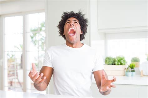 캐주얼 한 흰색 티셔츠를 입은 젊은 아프리카 계 미국인 남자가 집에 앉아 화가 나서 분노와 분노로 비명을 지르며 분노로 외쳤습니다 분노와 공격적인 개념 T 셔츠에 대한 스톡