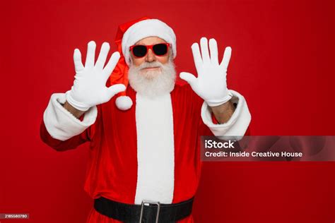 의상 모자 안경을 쓰고 카메라를 보며 손을 흔들며 행복한 재미 있는 시니어 늙은 수염 난 산타 클로스 빨간 배경 얼굴 사진 초상화에 고립 된 메리 크리스마스에 인사 12월에