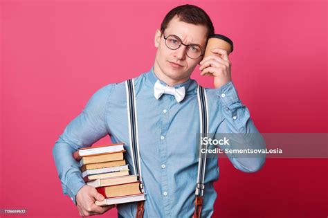 한 톤의 파란 셔츠에 있는 젊은 학생 졸린과 삼 출 한 외모 머리 근처에 커피를 보유 하 고 책의 거 대 한 스택 Axams에 대 한 Preparig에서 피곤 남성 장미 빛