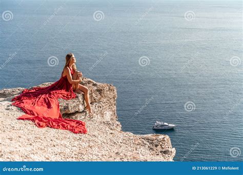 A Girl With Loose Hair In A Red Dress Sits On A Rock Rock Above The Sea