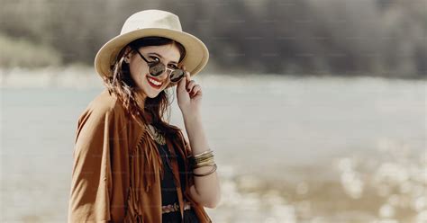 세련된 힙스터 보호 여자는 모자 가죽 가방 프린지 판초 및 액세서리로 선글라스를 끼고 웃고 있습니다 행복한 여행자 소녀 모습 산의 해변 근처 방랑벽 여름 여행