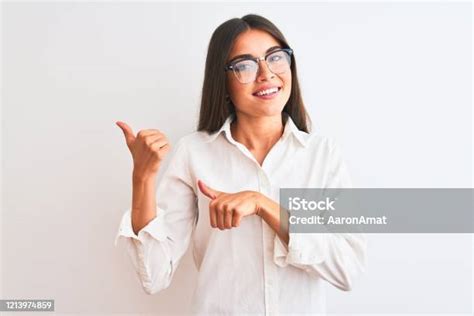고립된 흰색 배경 위에 안경을 쓰고 서있는 젊은 아름다운 사업가가 손과 엄지 손가락으로 뒤쪽을 가리키며 자신감을 가지고 미소짓고 있습니다 가리키기에 대한 스톡 사진 및 기타