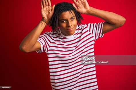고립 된 빨간 배경 위에 서있는 줄무늬 티셔츠를 입은 공포를 가진 젊은 아프로 남자는 손 손바닥으로 냉소적이고 회의적인 모습으로 토끼 귀 제스처를하고 있습니다 부활절 토끼