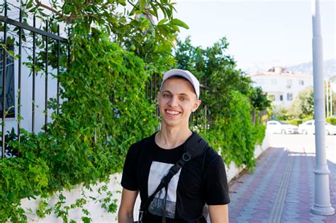 티셔츠와 모자를 쓴 웃는 남자가 잎이 무성한 보도에 서 있고 배경 녹지와 건물이 있습니다 20 24세에 대한 스톡 사진 및 기타 이미지 Istock