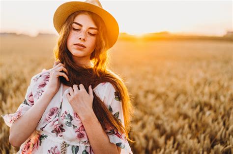 곱슬 머리와 주근깨 얼굴을 가진 아름다운 젊은 여성의 초상화입니다 일몰에 밀 밭에서 포즈를 취하고 카메라를 보고 있는 드레스와 모자를 입은 여성 갈색 머리에 대한 스톡 사진