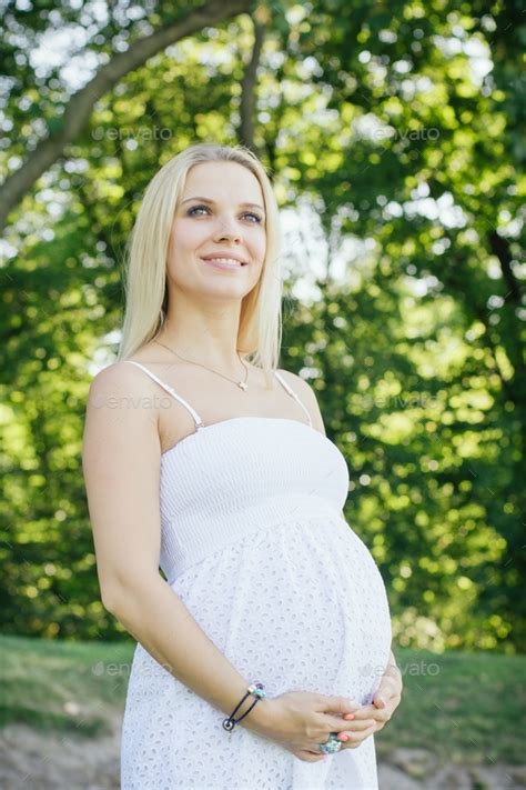 Pregnant Smiling Blond Woman In White Dress Looking Up And Holding Her Big Belly In The Park