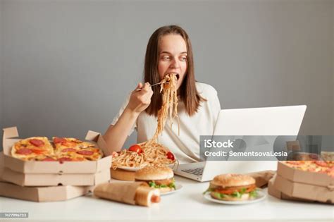 노트북에서 작업하는 동안 패스트 푸드를 먹는 바쁜 배고픈 여성의 실내 장면 회색 벽에 기대어 테이블에 앉아 노트북에서 작업하고 파스타를 먹는 것은 입을 벌리고 노트북