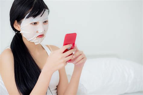 침실에서 침대에서 스마트 폰을보고 앉아 시트 얼굴 마스크를 가진 아름다운 젊은 아시아 여성 집에서 주름의 스킨 케어에 대한 얼굴 마스크를 적용 하는 뷰티 걸 피부 관리 건강